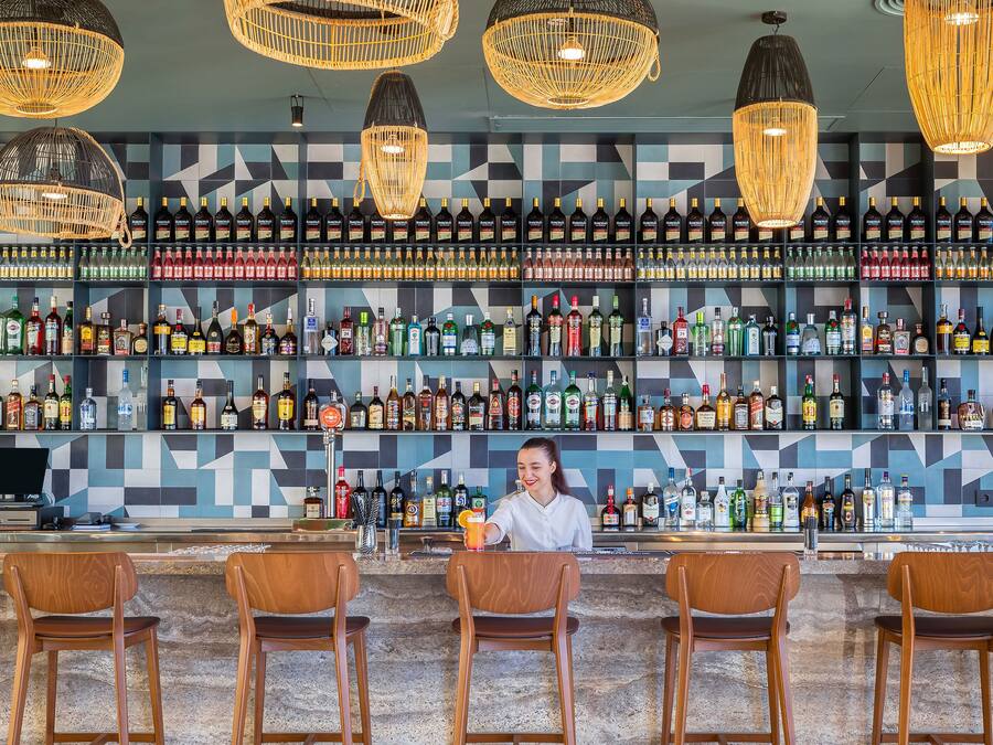 Uma área de bar com várias prateleiras de garrafas de bebidas, um barman e três banquetas de madeira.