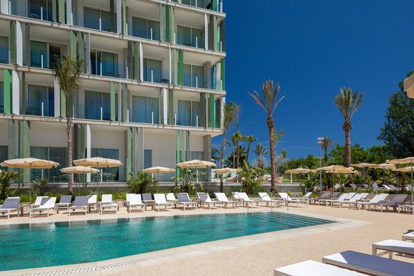 Área de piscina de hotel com espreguiçadeiras e guarda-sóis, cercada por palmeiras e um edifício com varandas.