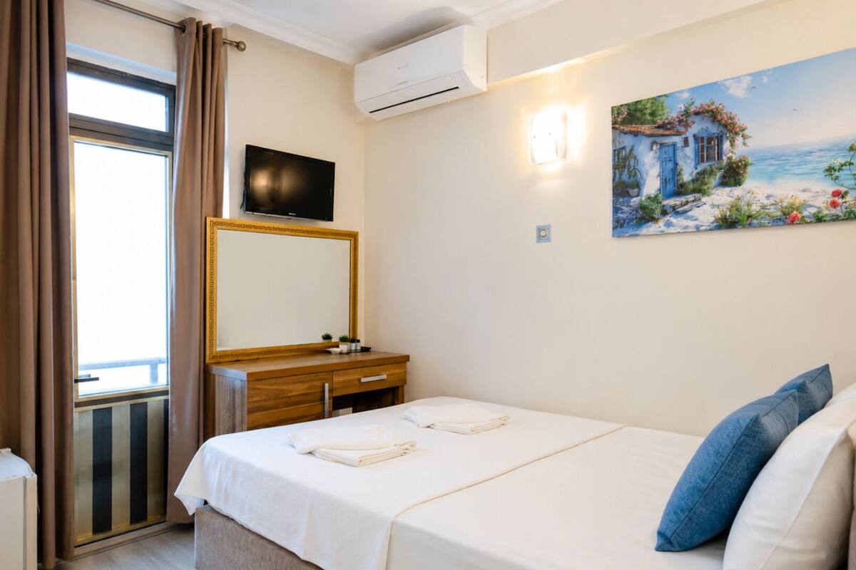 Quarto de hotel com cama, televisão, quadro, janela com cortinas e aparelho de ar condicionado.