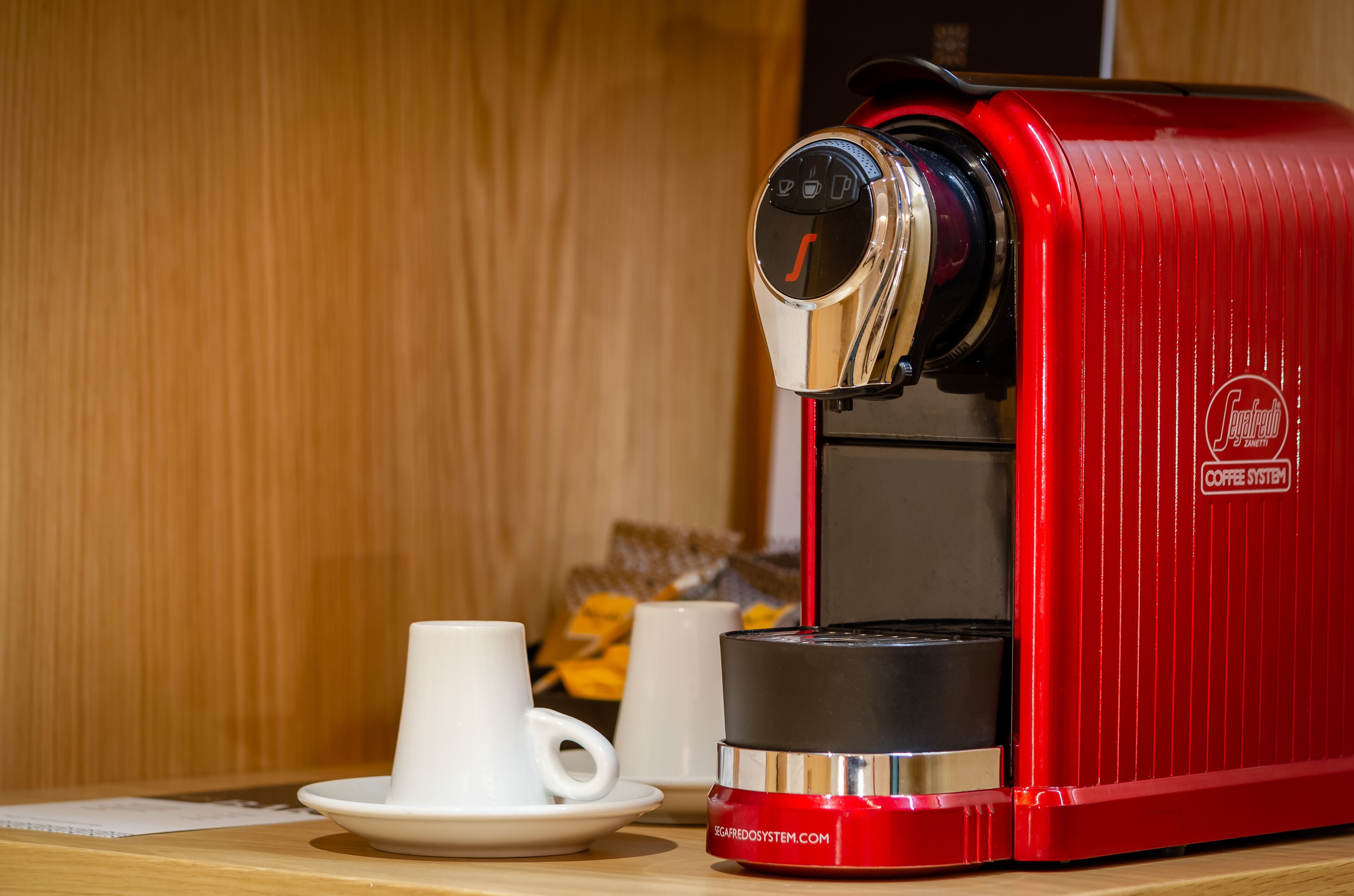 Uma máquina de café vermelha com visor digital, uma chávena branca e um prato sobre uma mesa de madeira.