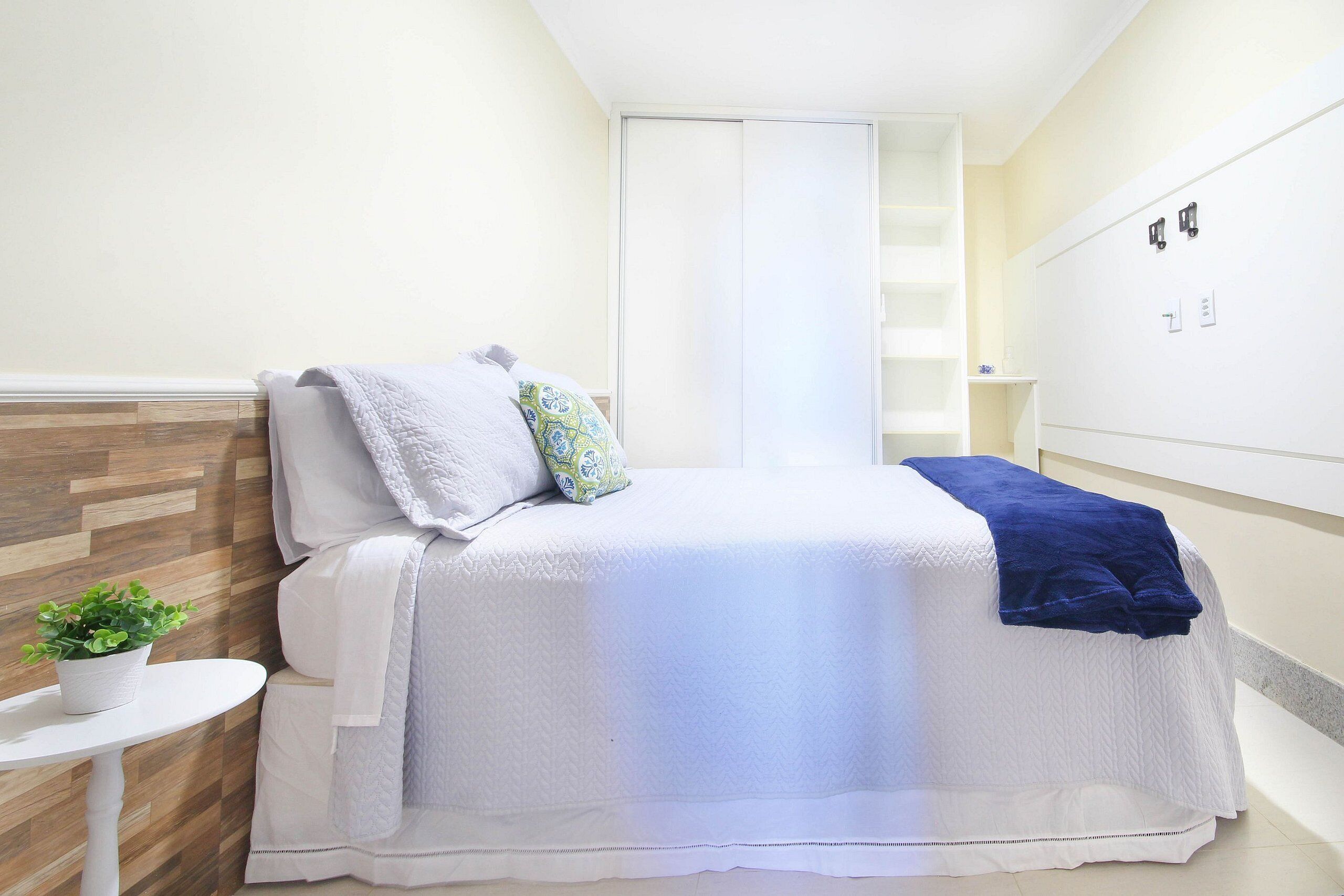 Quarto bem arrumado com uma cama, almofadas, uma mesinha com uma planta e um guarda-roupa branco.