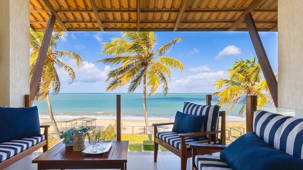Um lounge à beira-mar com vista para o oceano, com sofás listrados e uma mesa de madeira com bandeja de vidro.