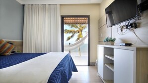 Quarto de hotel com uma cama, uma televisão na parede, uma escrivaninha com uma planta e vista para a praia através de uma porta.