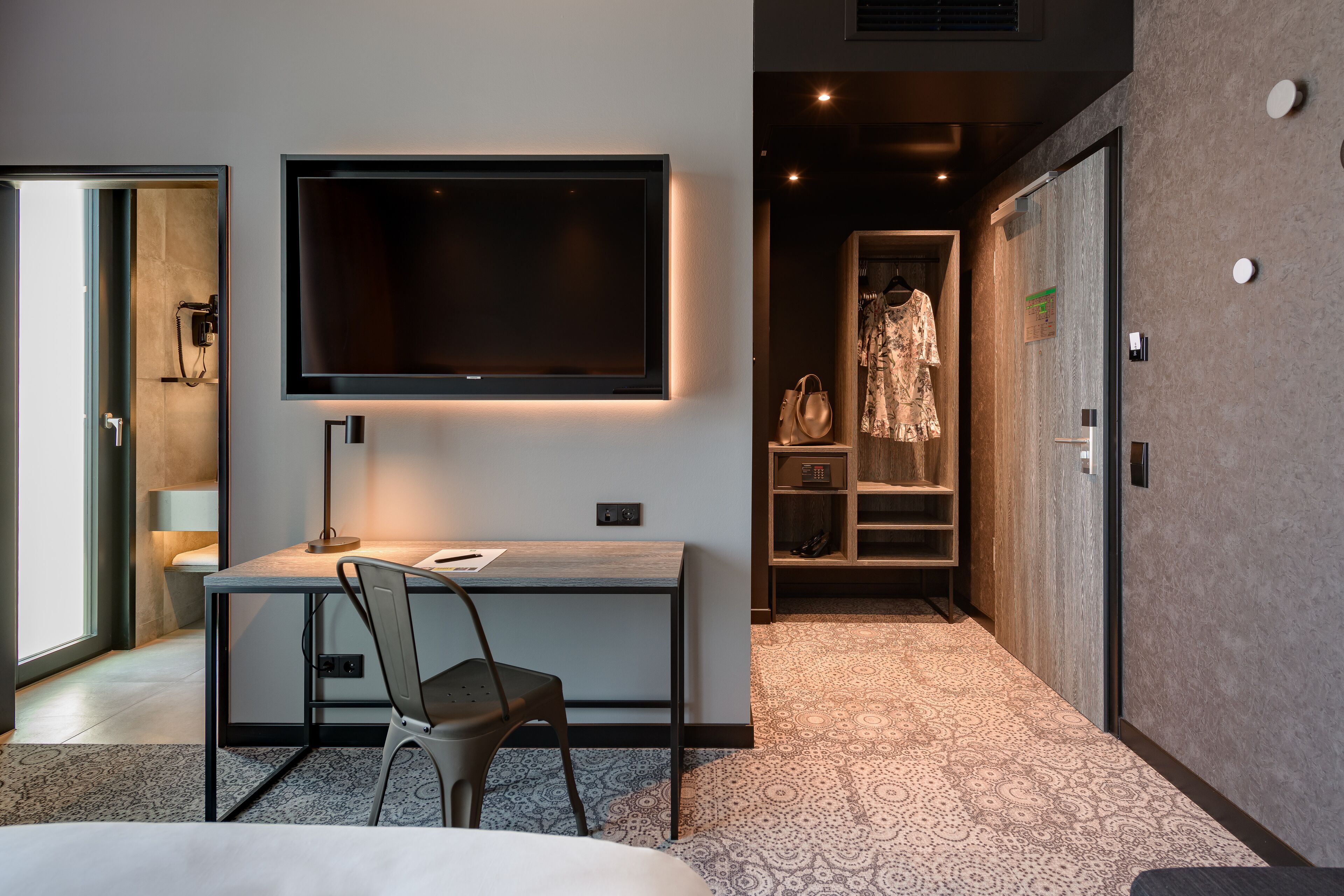 Quarto de hotel com escrivaninha, cadeira, televisão, guarda-roupa e carpete estampado.