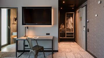 Quarto de hotel com escrivaninha, cadeira, televisão, guarda-roupa e carpete estampado.