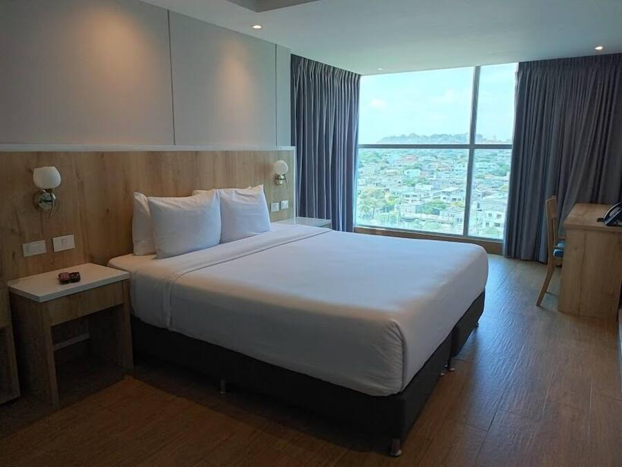 Quarto de hotel moderno com uma cama grande, cabeceira de madeira, mesas de cabeceira e vista para a paisagem urbana através de amplas janelas.