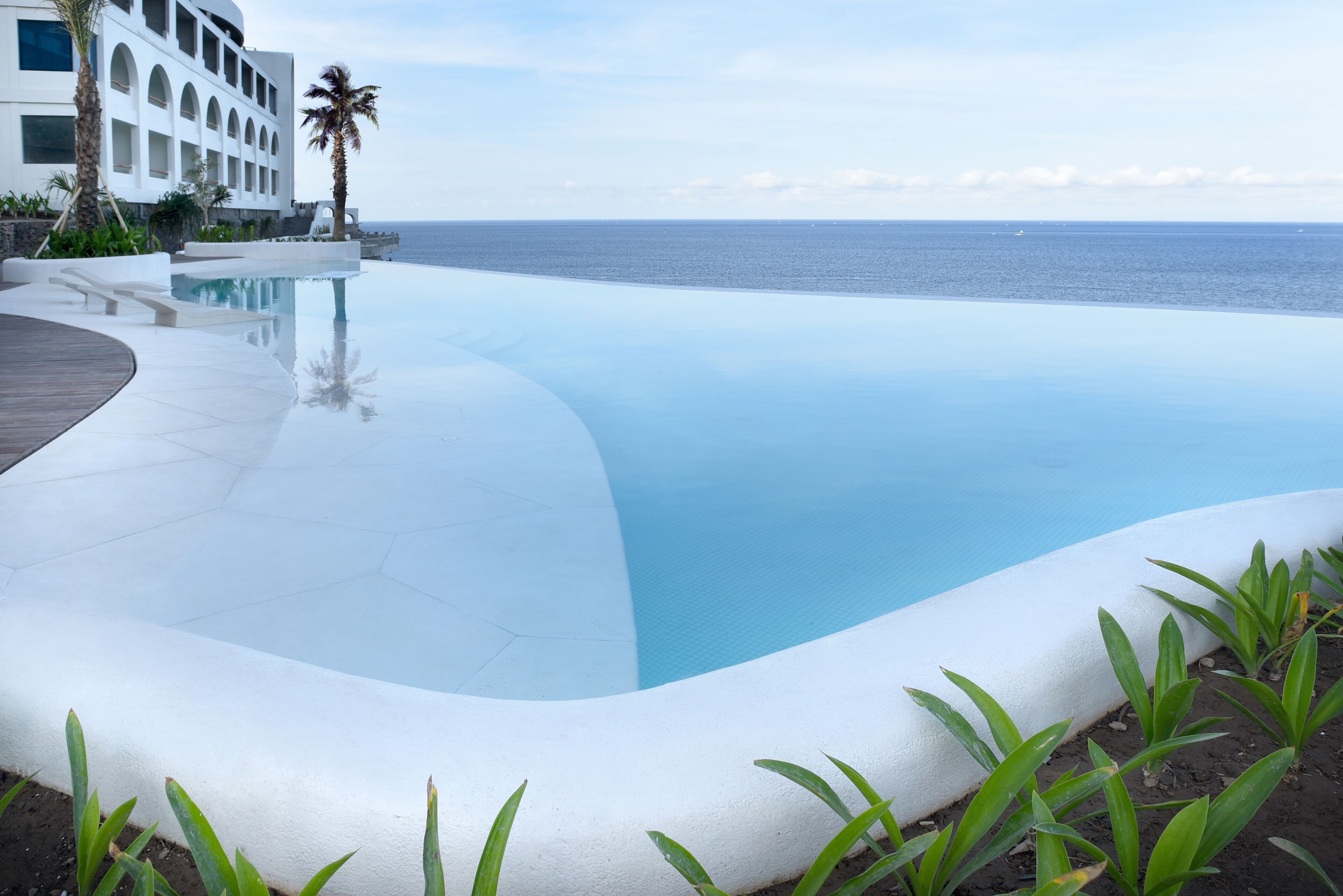 Piscina infinita moderna com vista para o oceano, rodeada por um deck branco e palmeiras.