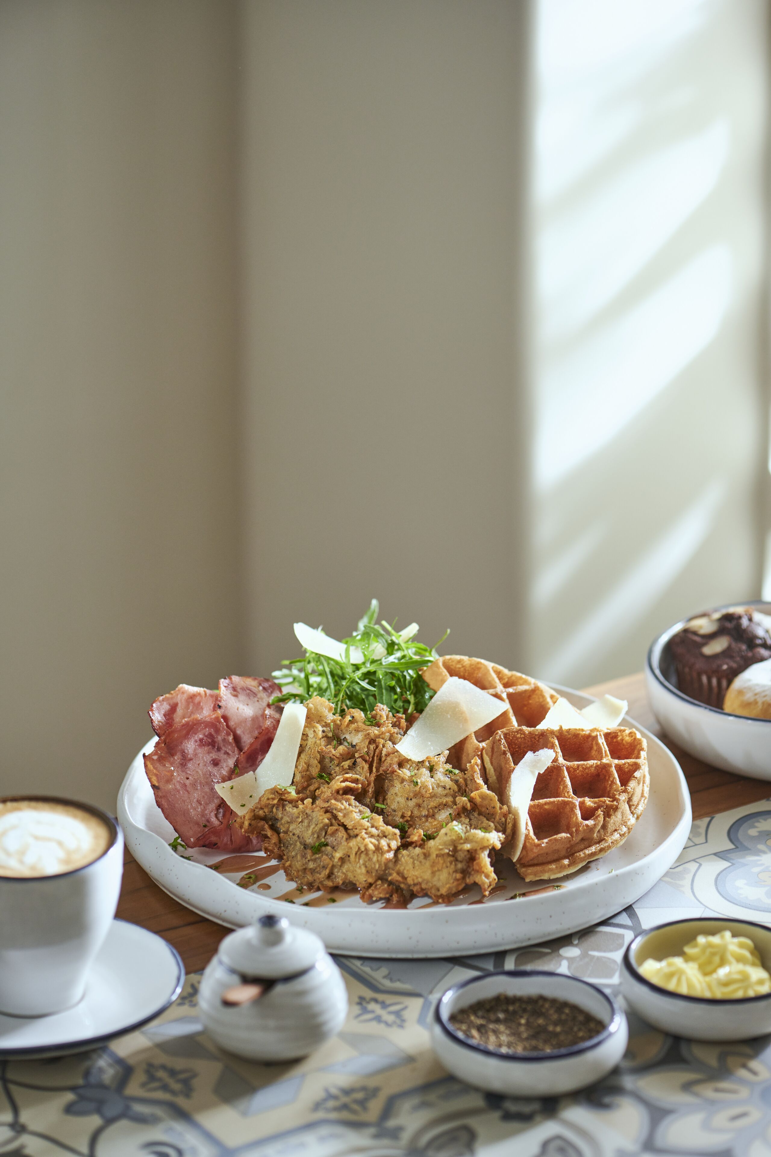 Prato de comida com frango frito, waffles e bacon, acompanhado de condimentos e uma xícara de café sobre uma mesa.