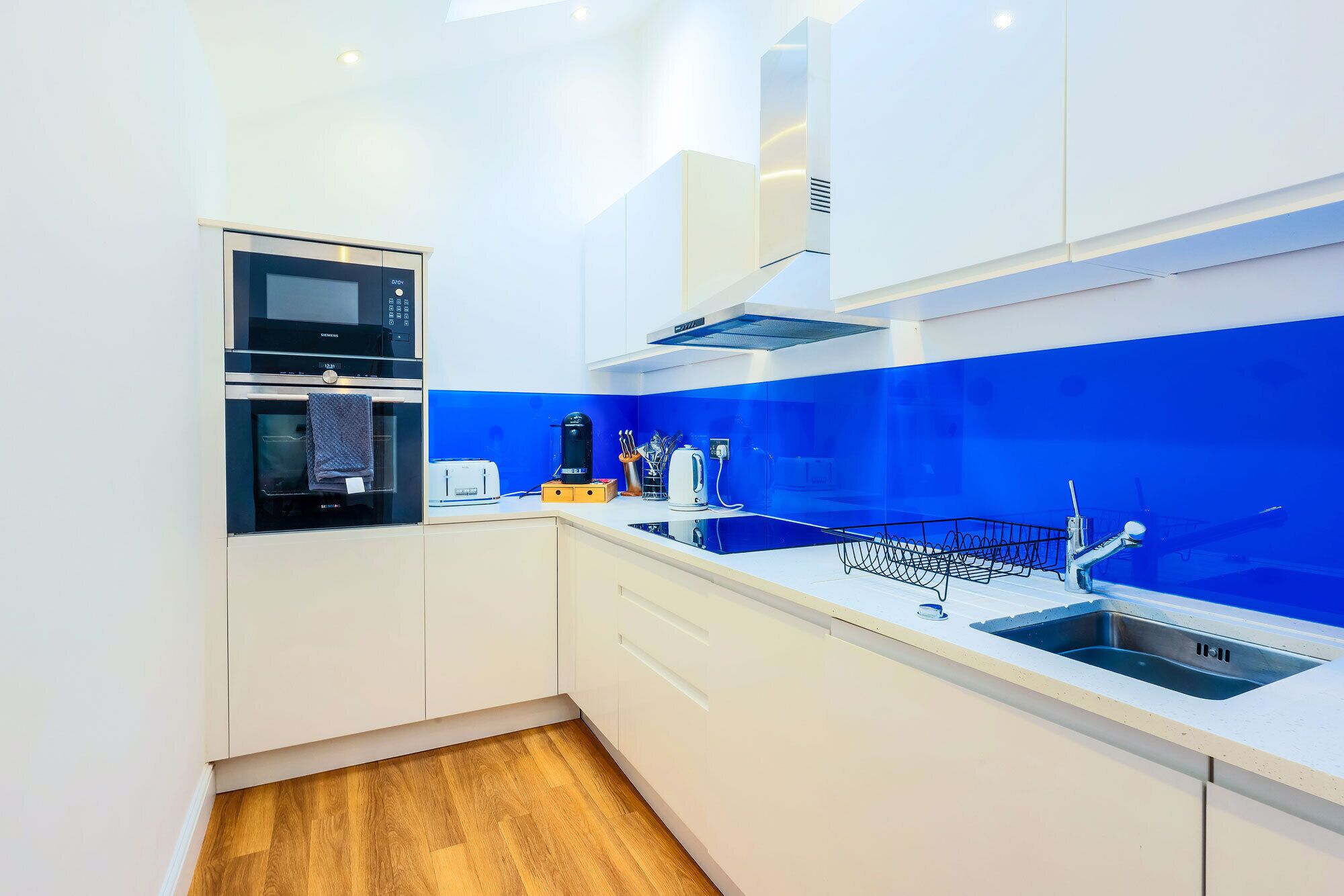 Uma cozinha moderna com armários brancos, painel de parede azul, forno embutido, pia e chaleira.