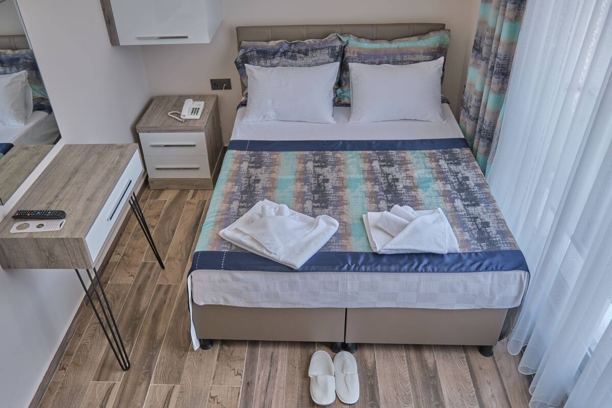 Uma cama bem arrumada com um cobre-leito estampado, um telefone de apoio e um par de chinelos no chão.