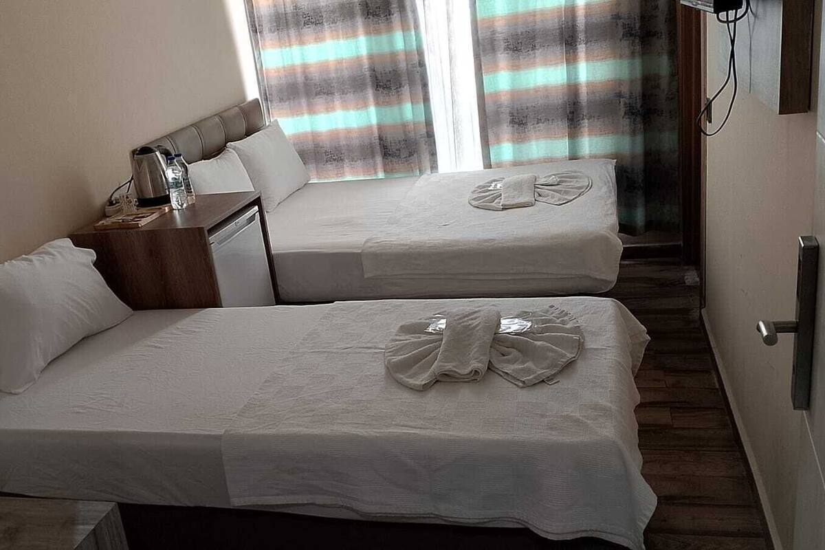 Quarto de hotel com duas camas, uma mesa de cabeceira com abajur e uma janela com cortinas listradas.