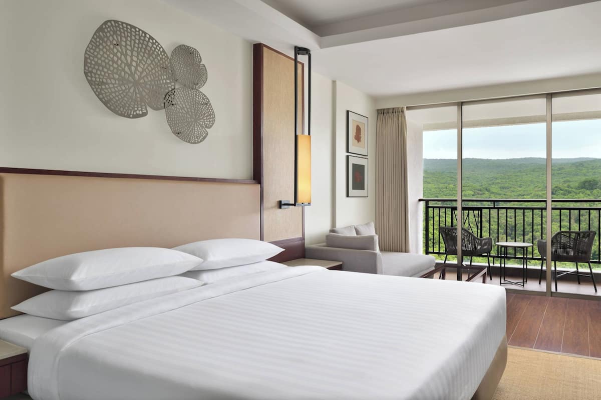 Quarto de hotel com uma cama grande, área de estar com mesa e cadeiras, e varanda com vista para o verde.