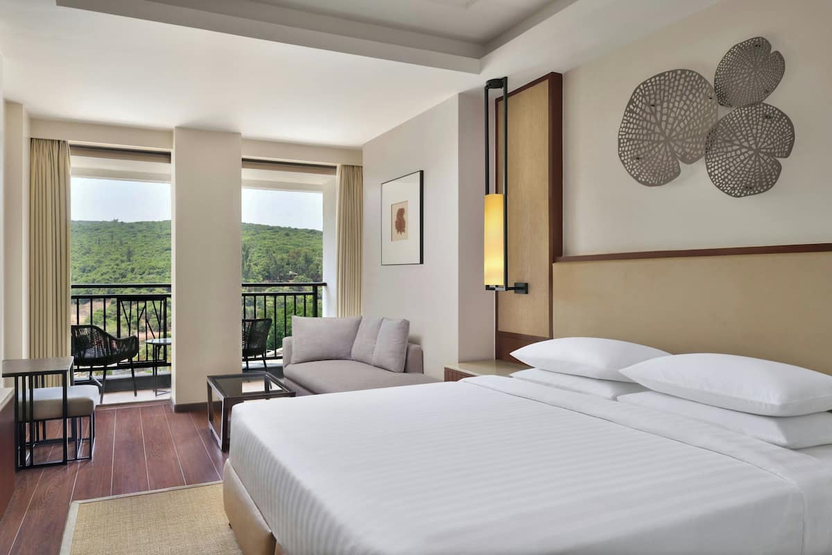 Quarto de hotel com uma cama grande, um sofá, uma mesa pequena e uma varanda com vista para o verde.