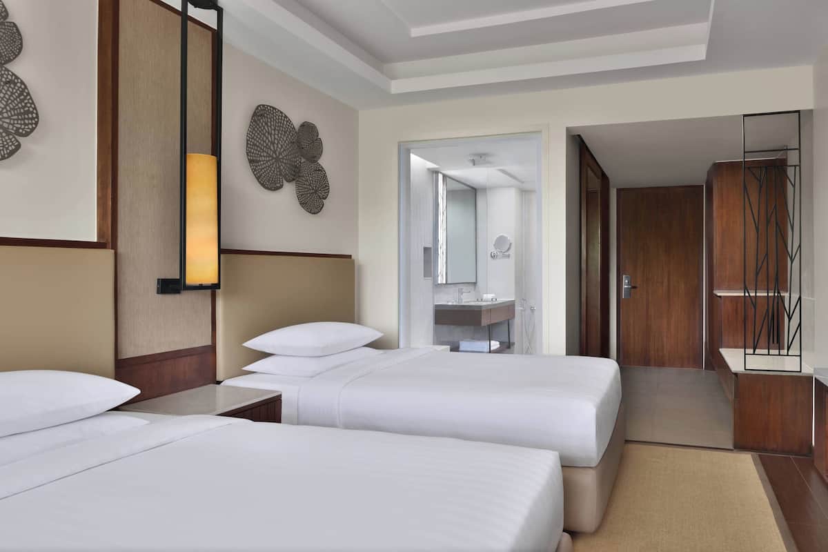 Quarto de hotel com duas camas, um espelho grande, banheiro com pia e um guarda-roupa de madeira.