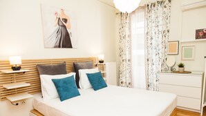 Quarto bem arrumado com cabeceira de madeira, duas mesas de cabeceira com abajures, cama com roupa de cama branca e azul, e cortina florida.