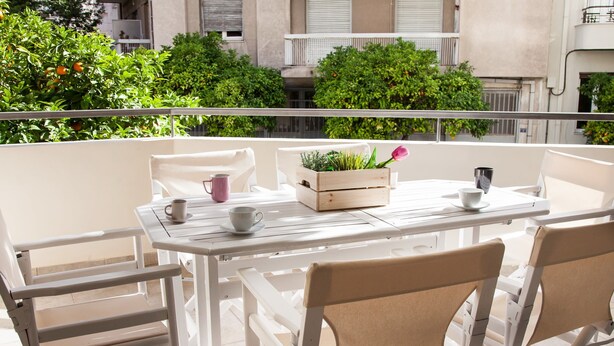 Uma varanda com mesa e cadeiras brancas, um floreiro de madeira com plantas e uma flor rosa, e vista para árvores verdes e edifícios.