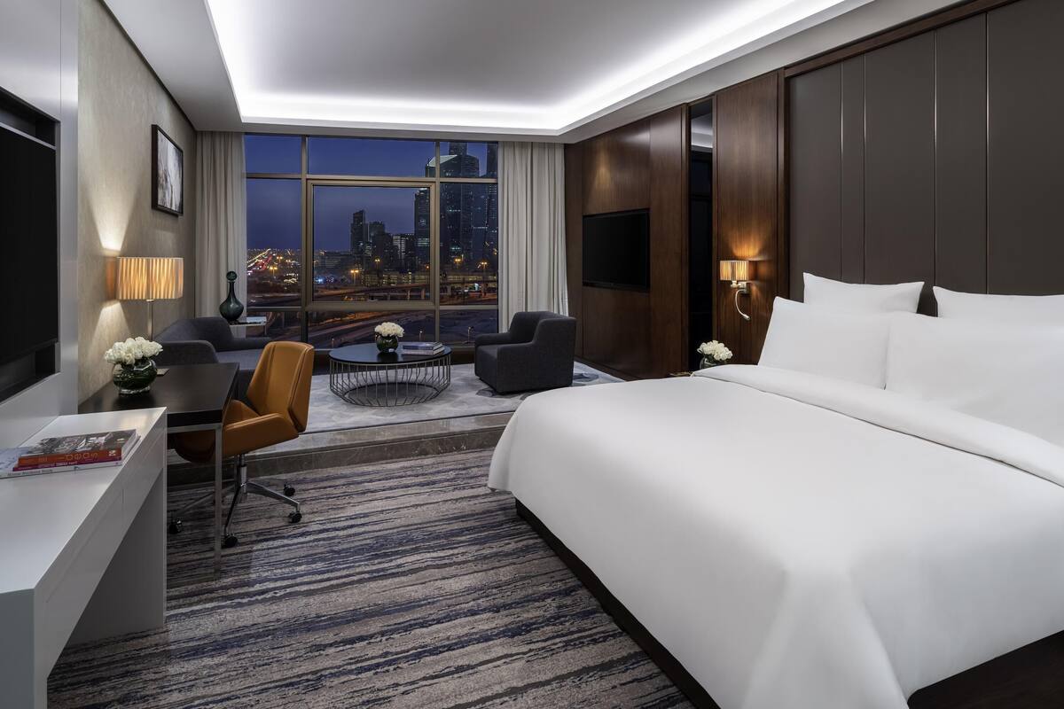 Quarto de hotel com cama grande, escrivaninha, cadeira, mesa de centro e vista da paisagem urbana à noite.
