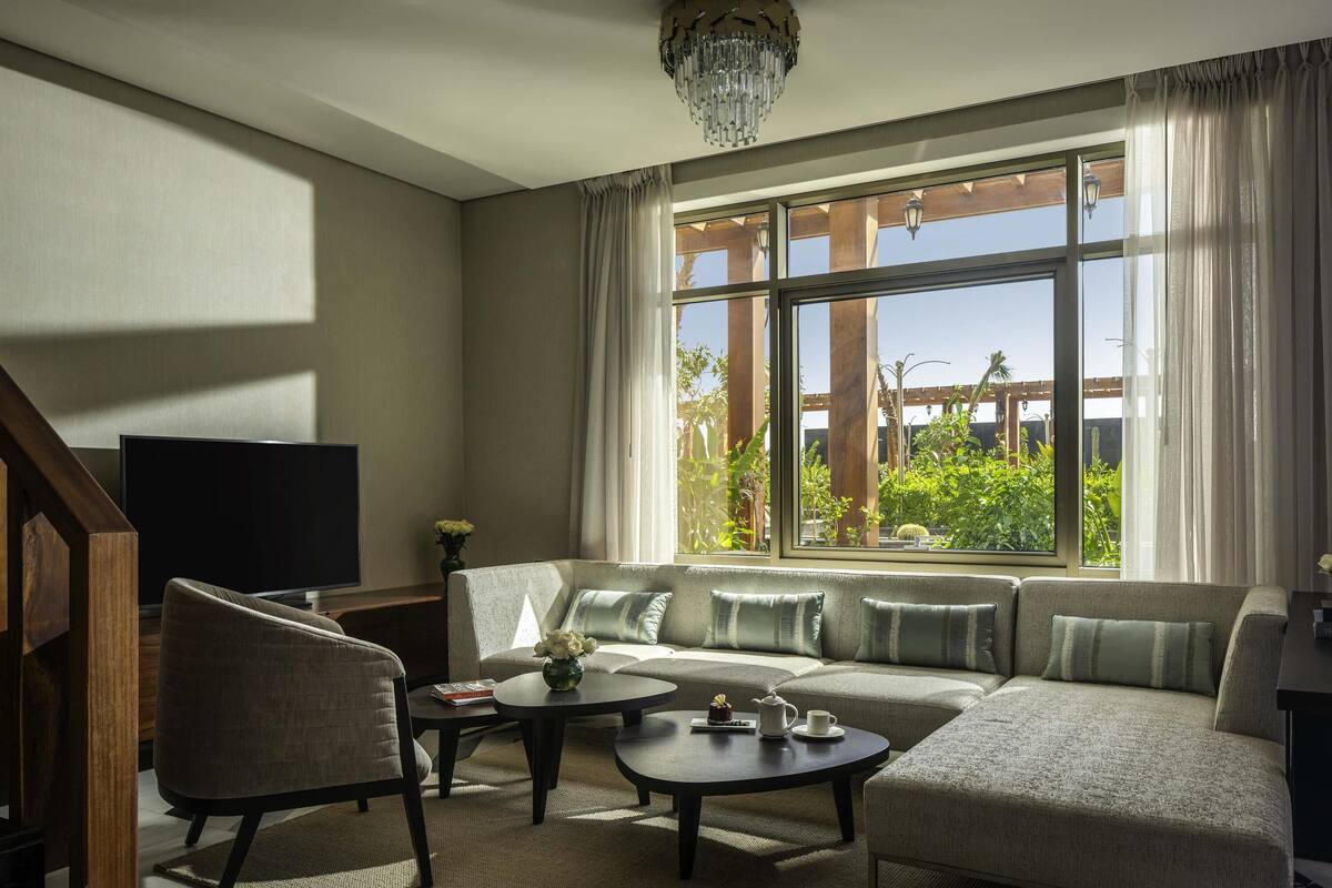 Uma sala de estar moderna com sofá, poltronas e uma mesa de centro. Uma televisão está fixada na parede, e há uma janela grande com cortinas.