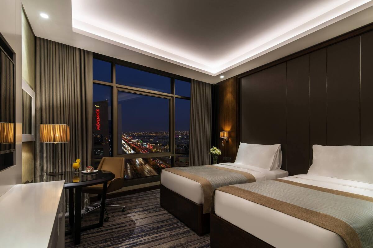 Quarto de hotel com duas camas, uma escrivaninha, uma cadeira e uma janela grande com vista para a cidade.