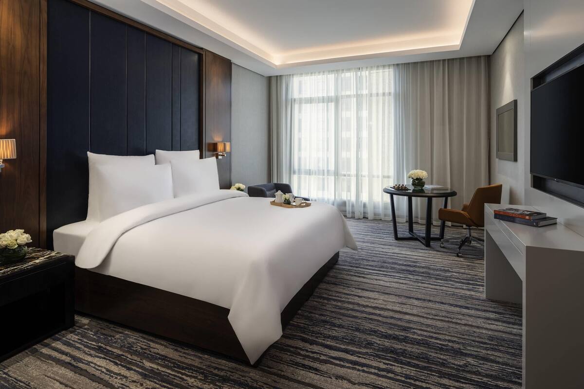 Quarto de hotel com uma cama grande, uma escrivaninha, uma cadeira, uma televisão e uma janela com cortinas.