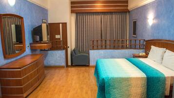 Quarto de hotel com cômoda de madeira, televisão, cama com colcha verde-água, mesa de cabeceira e uma cadeira.