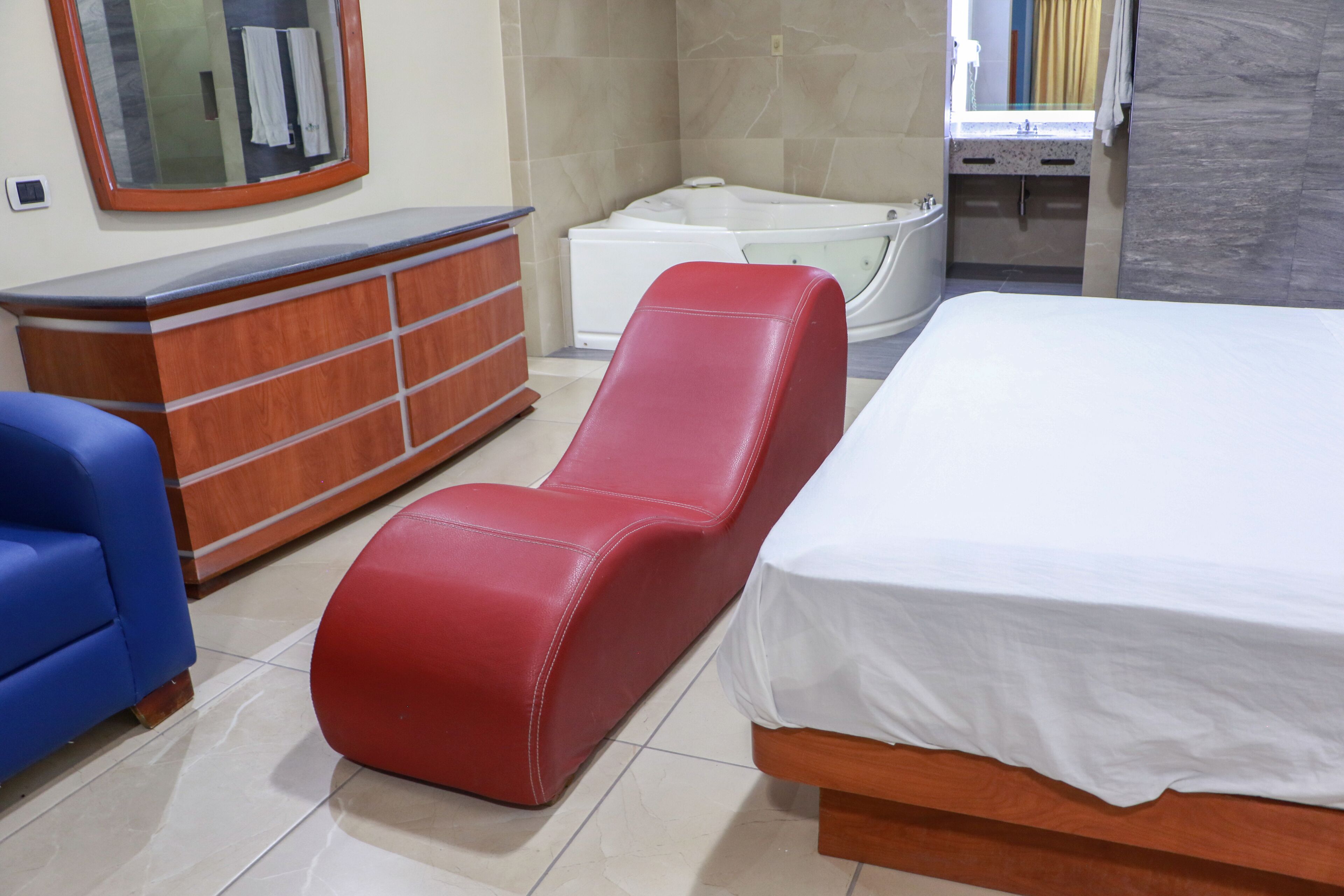 Quarto de hotel com cama, poltrona reclinável vermelha, penteadeira de madeira, poltrona azul, banheira e espelho.