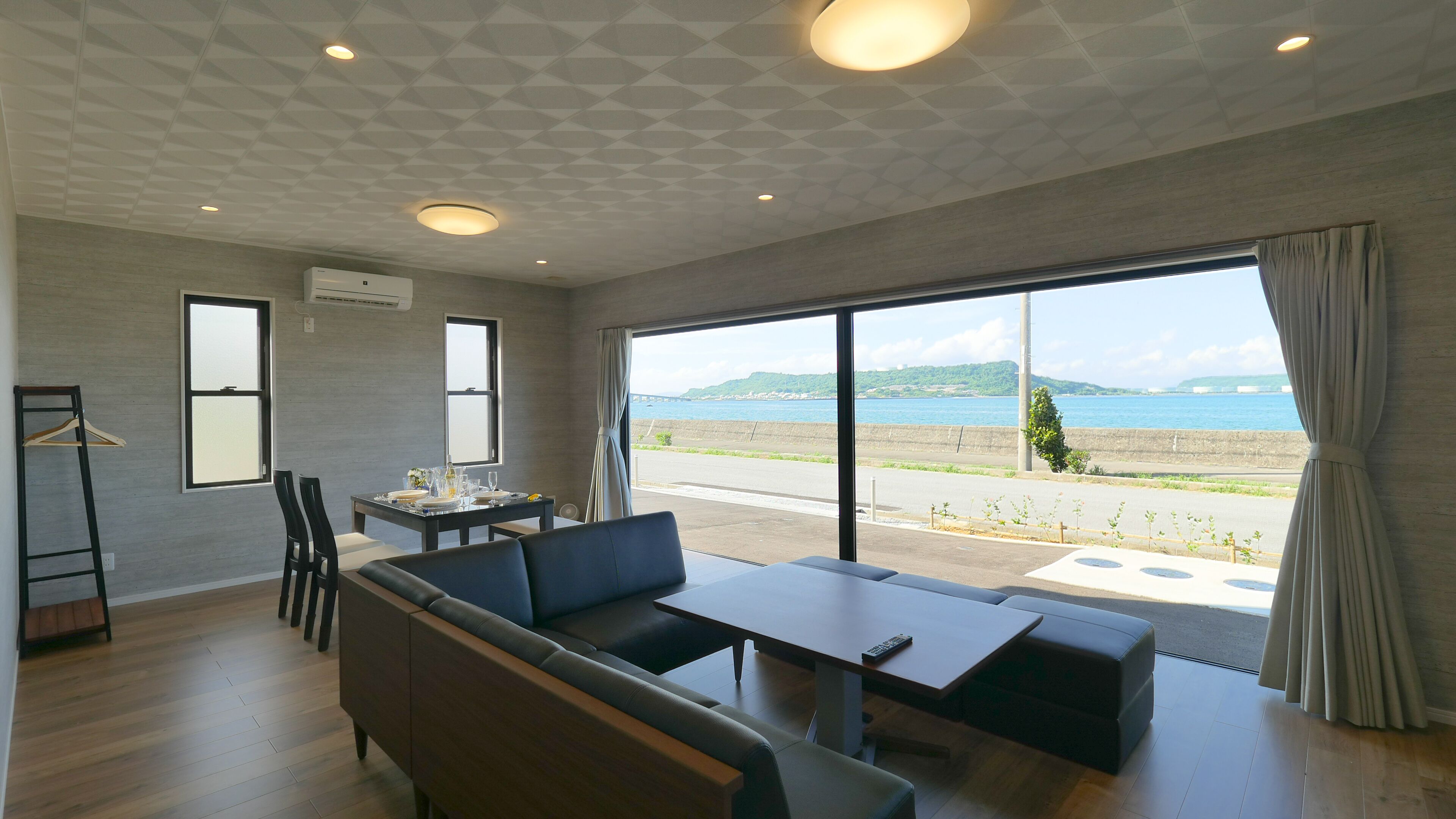 Quarto moderno com mesa de jantar, sofá e grandes janelas com vista para o mar.
