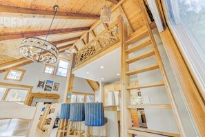 Interior em madeira com teto alto, área de refeições com cadeiras listradas e cozinha com eletrodomésticos modernos.