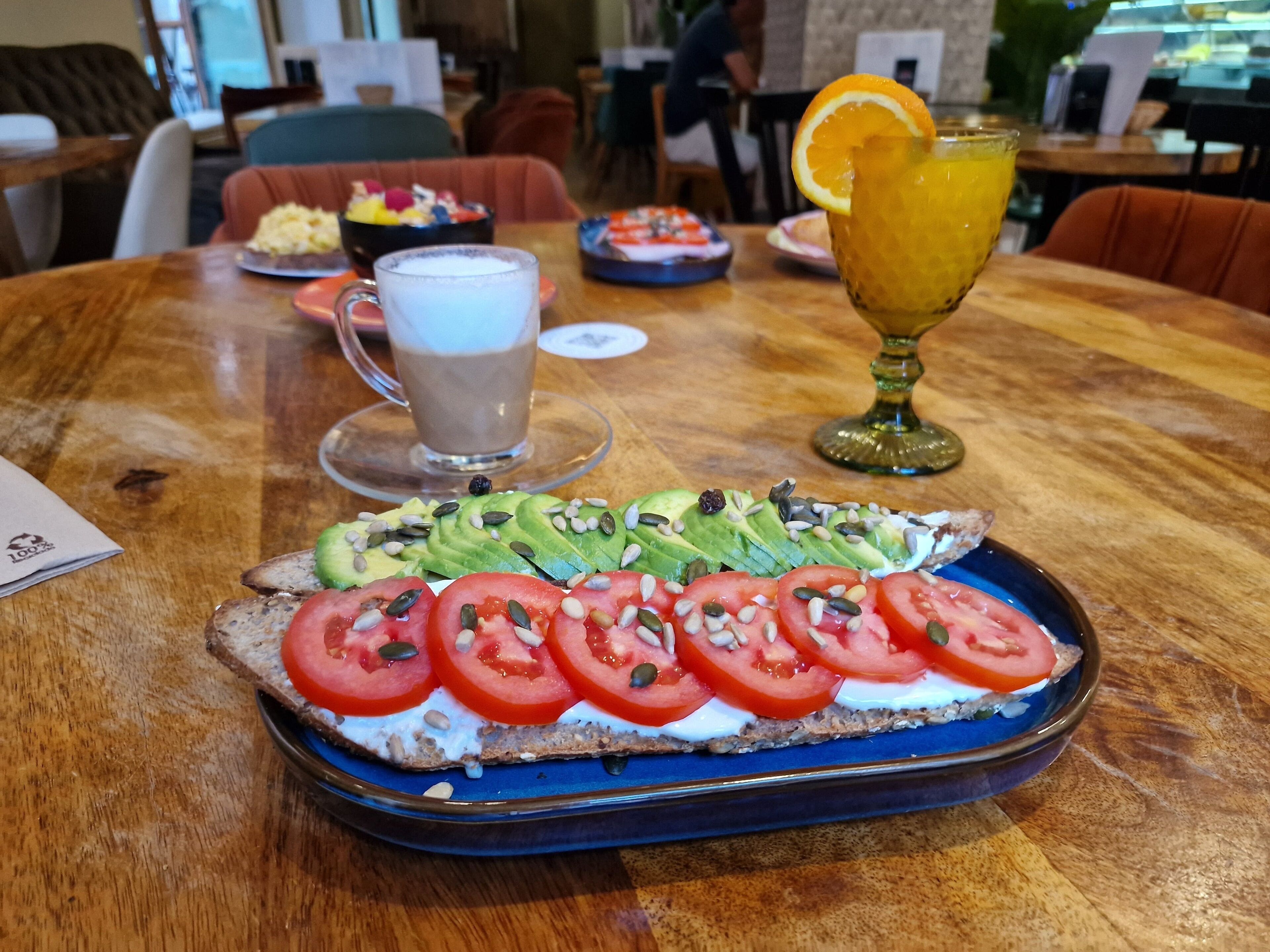 Uma refeição com fatias de abacate, tomate e sementes em um prato, um copo de café e um copo de suco de laranja sobre uma mesa de madeira.