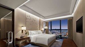 Quarto de hotel moderno com uma cama grande, abajures, televisão de tela plana, vista para a cidade através de janelas amplas e um espelho em uma parede.