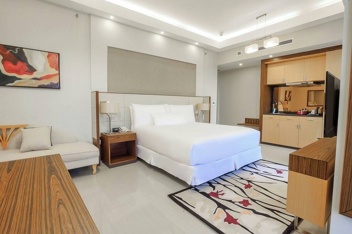 Quarto de hotel moderno com uma cama grande, uma escrivaninha, uma cadeira, uma mesa de cabeceira, uma televisão e uma cozinha compacta.