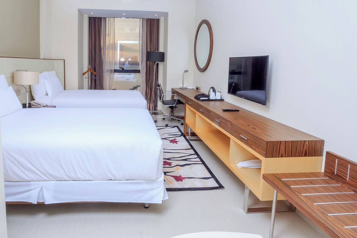 Quarto de hotel com duas camas, uma mesa, uma televisão e vista para o exterior.