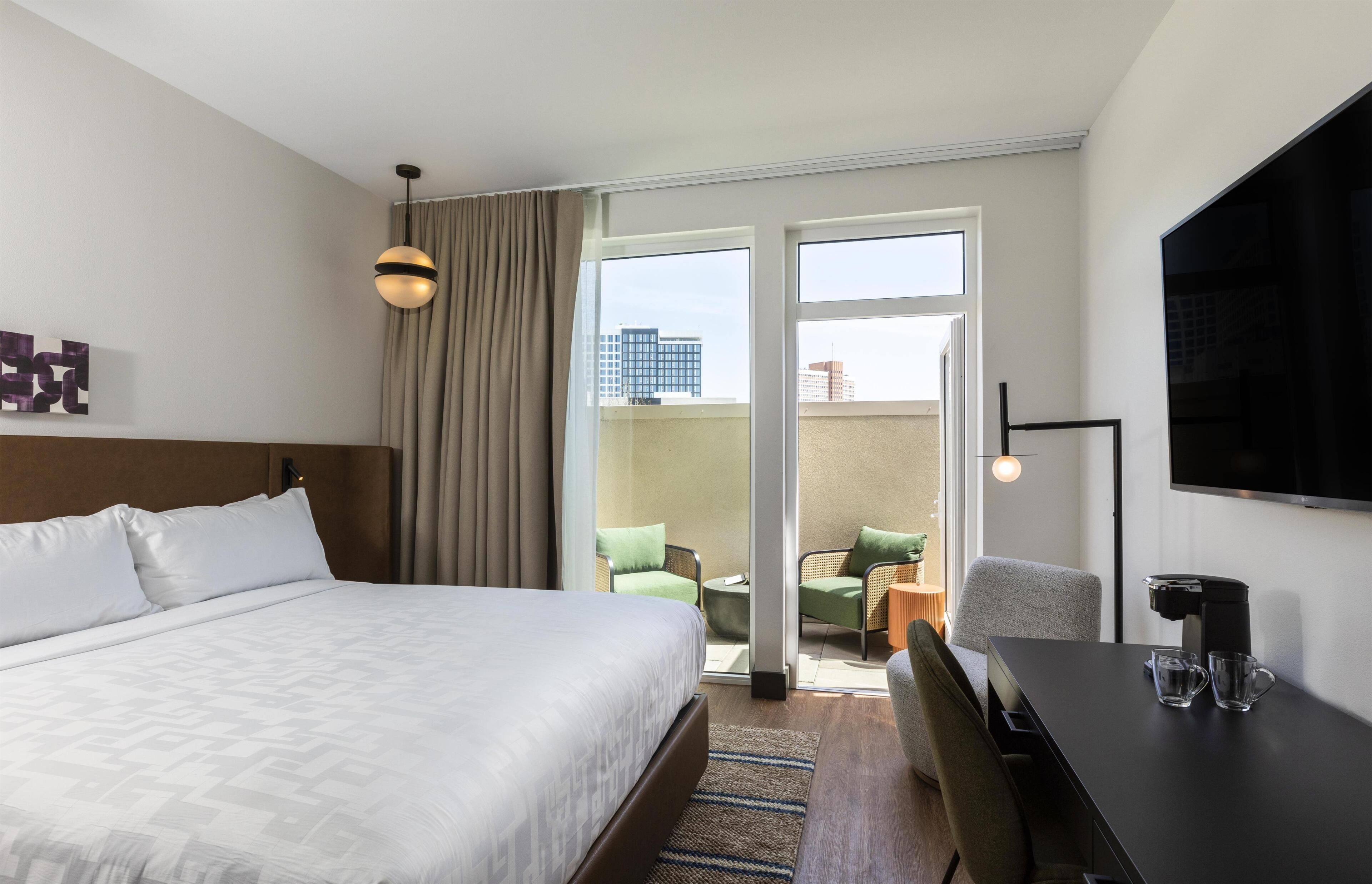 Quarto de hotel com cama, escrivaninha, cadeira, televisão e varanda com cadeiras.