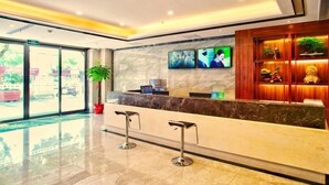 Um lobby de hotel moderno com um balcão de recepção, dois banquetas de bar, uma grande planta em vaso e uma televisão na parede.