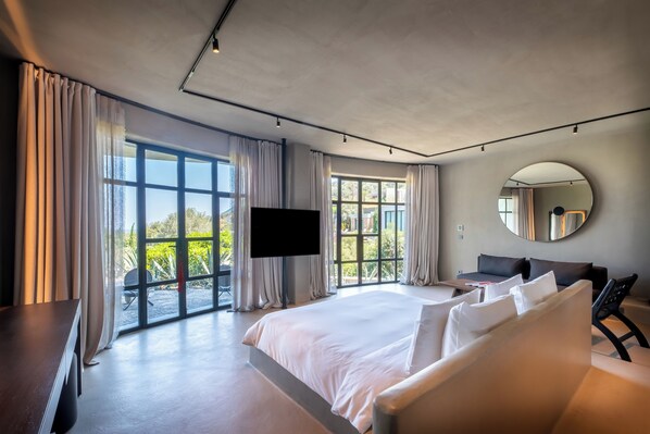 Quarto de hotel moderno com uma cama grande, uma mesa de trabalho e uma televisão. Há grandes janelas e vista para o verde lá fora.