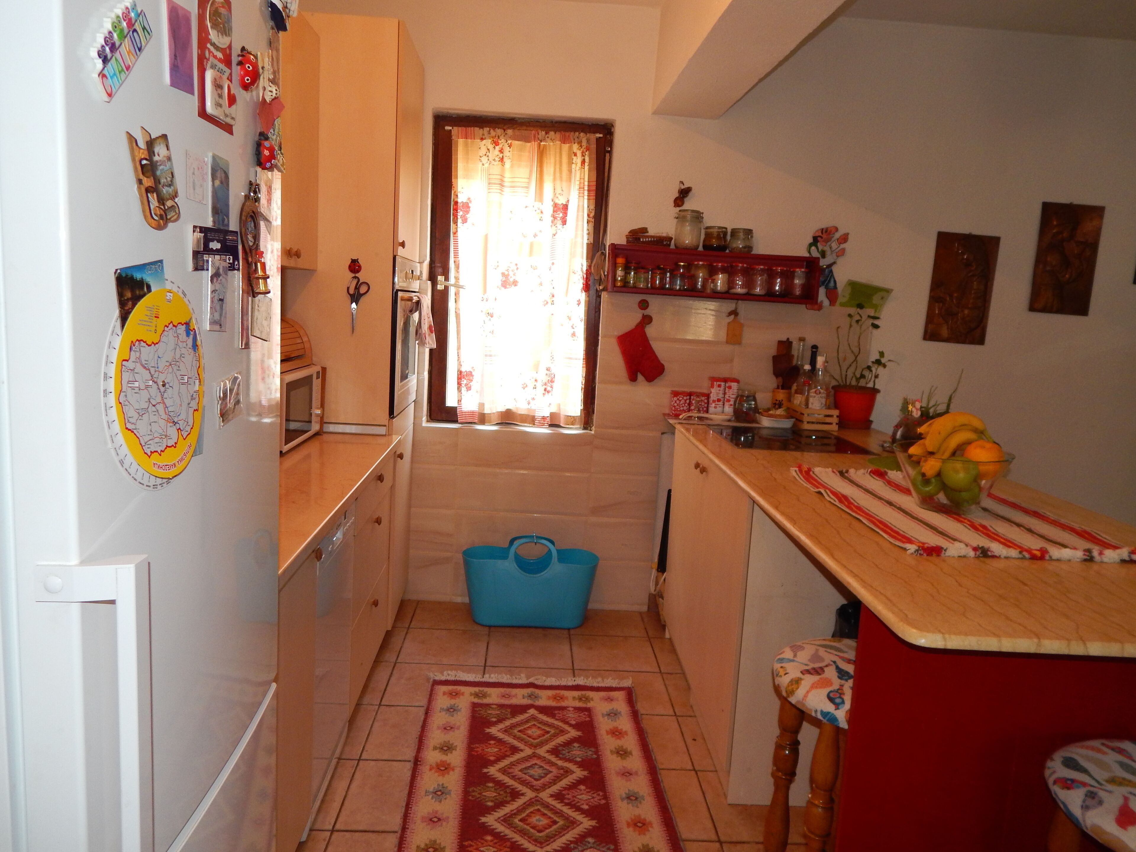 Uma cozinha compacta com micro-ondas, forno e uma pia pequena. Há uma janela com cortinas, uma lixeira azul e uma fruteira no balcão.