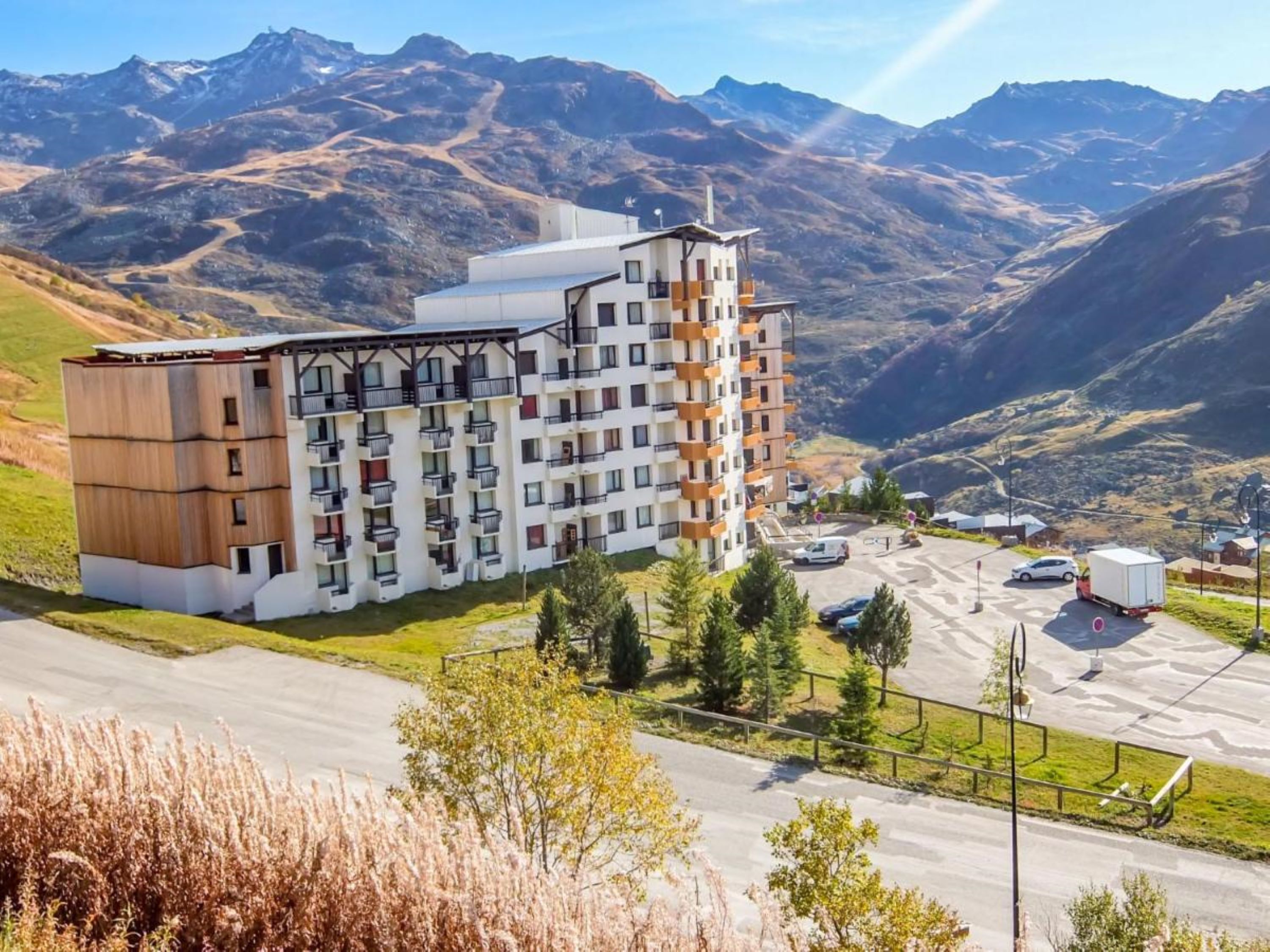 Hotel com vários andares, varandas, área de estacionamento e montanhas ao fundo.