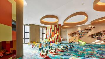 Um espaço interior bem iluminado, com designs geométricos coloridos nas paredes, área de recreação com brinquedos e um chão vibrante, e escada com estantes embutidas.