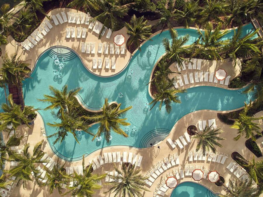 Vista aérea de uma piscina de resort com várias áreas de solário e palmeiras.
