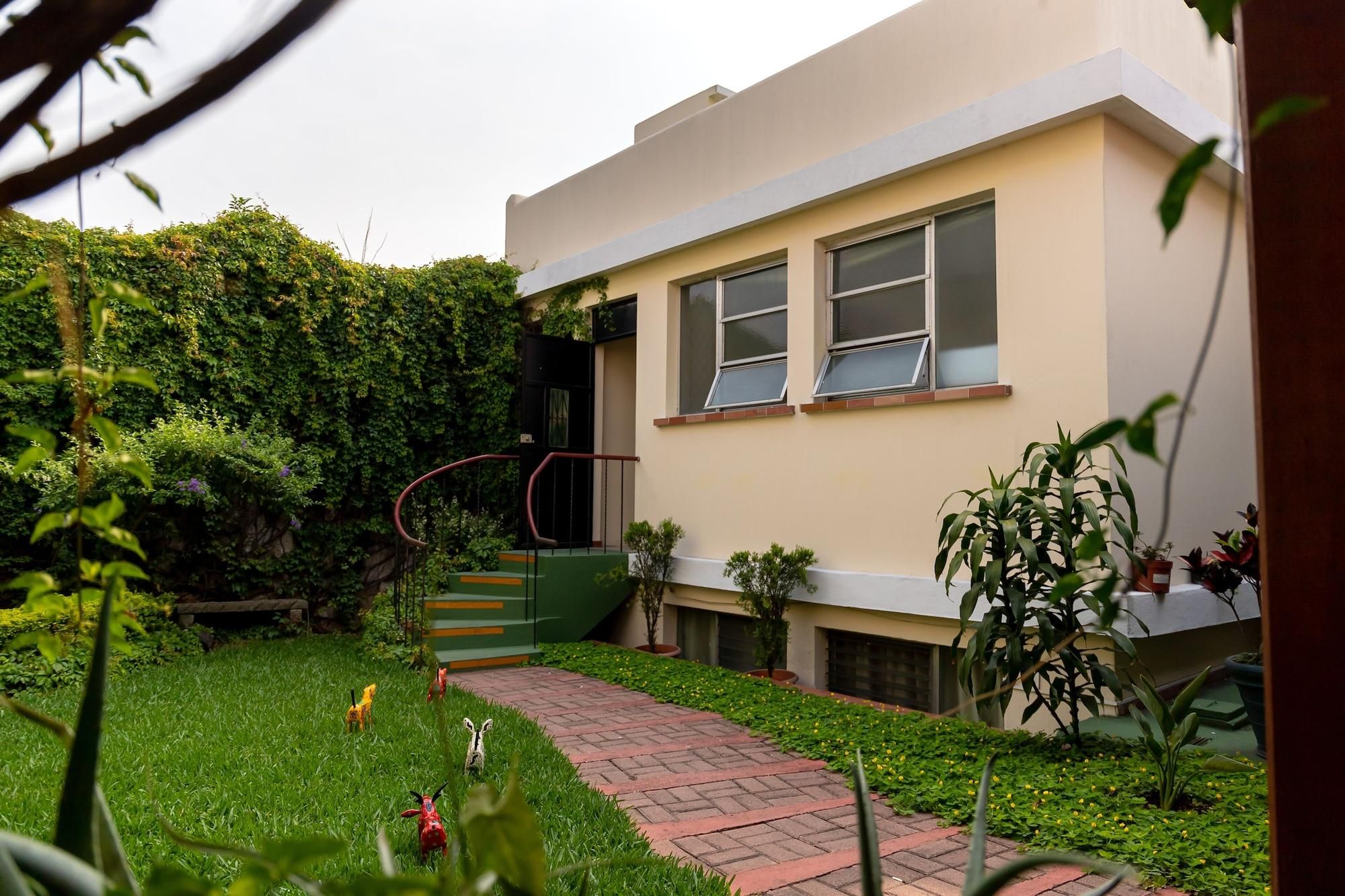 Uma casa moderna de dois andares com um caminho pavimentado que leva a uma porta verde, cercada por vegetação exuberante e plantas pequenas.