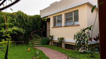 Uma casa moderna de dois andares com um caminho pavimentado que leva a uma porta verde, cercada por vegetação exuberante e plantas pequenas.