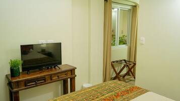 Quarto com cama, televisão sobre suporte de madeira, janela com cortinas e pequena mesa com planta.