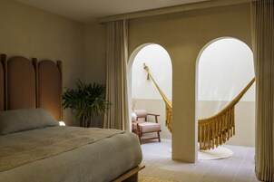 Quarto com cama, planta em vaso e vista através de uma porta arqueada para uma varanda com rede.