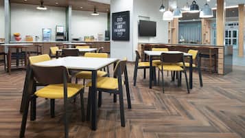 Uma sala de jantar moderna com piso de madeira, cadeiras amarelas e mesas. Há um balcão com um menu e uma televisão na parede.