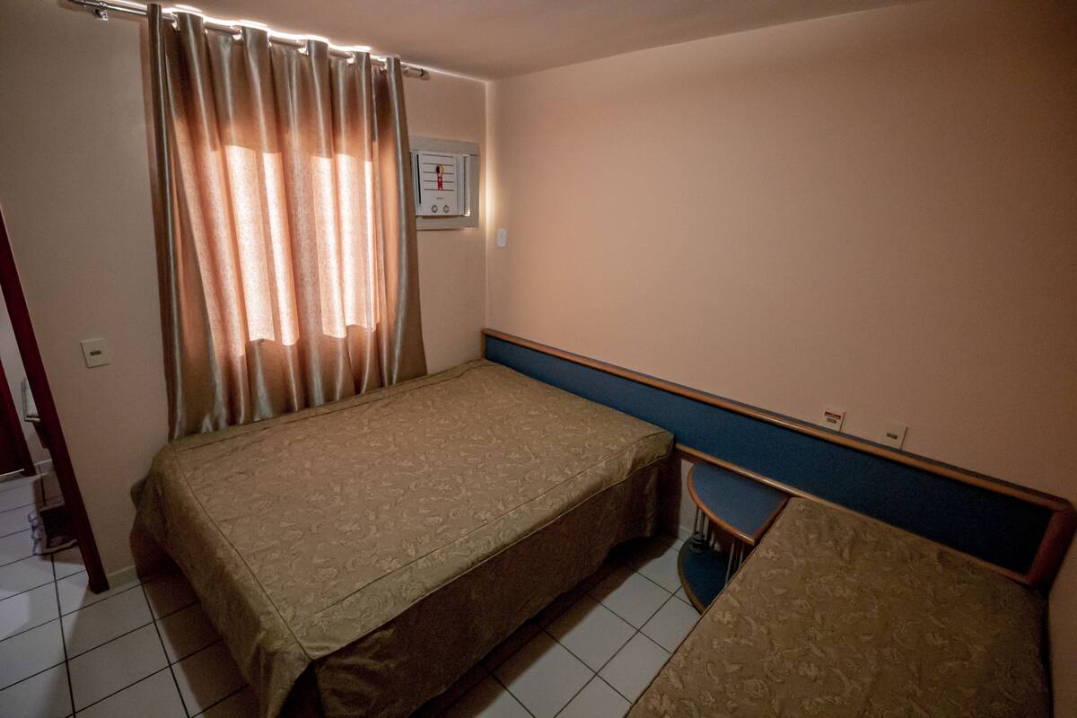 Quarto pequeno com uma cama de solteiro, cabeceira de madeira, mesa de cabeceira e janela com cortinas.