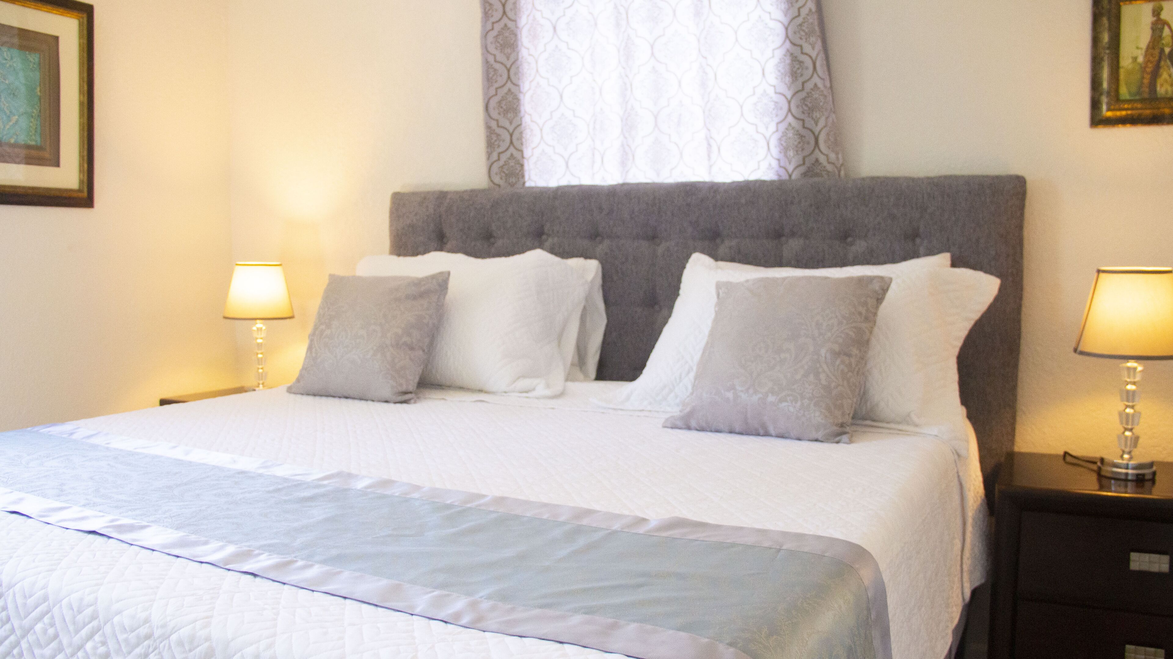 Uma cama bem arrumada com cabeceira cinzenta, roupa de cama branca e duas luminárias de cabeceira.