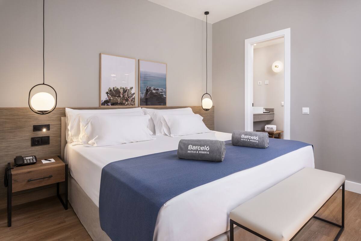 Quarto de hotel com uma cama grande, mesas de cabeceira, um banco e obras de arte na parede.