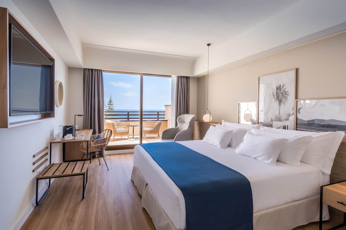 Quarto de hotel moderno com uma cama grande, uma escrivaninha e uma varanda com vista para o oceano.
