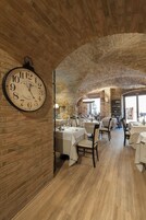 Restaurante com paredes de tijolos, um relógio grande e mesas postas para refeições.