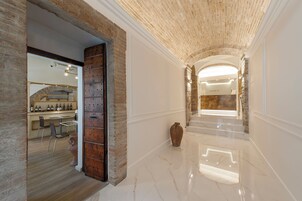 Corredor de hotel com parede de tijolos, um armário de madeira, piso de mármore e vista para um quarto com cama e banheiro.